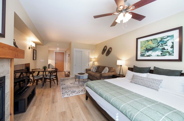 Quarto com cama, ventoinha no teto, lareira, área de refeições com mesa e cadeiras e corredor que dá para outro cômodo.