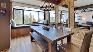 Uma área de refeições com mesa e bancos de madeira, janelas amplas com vista para árvores e um lustre.