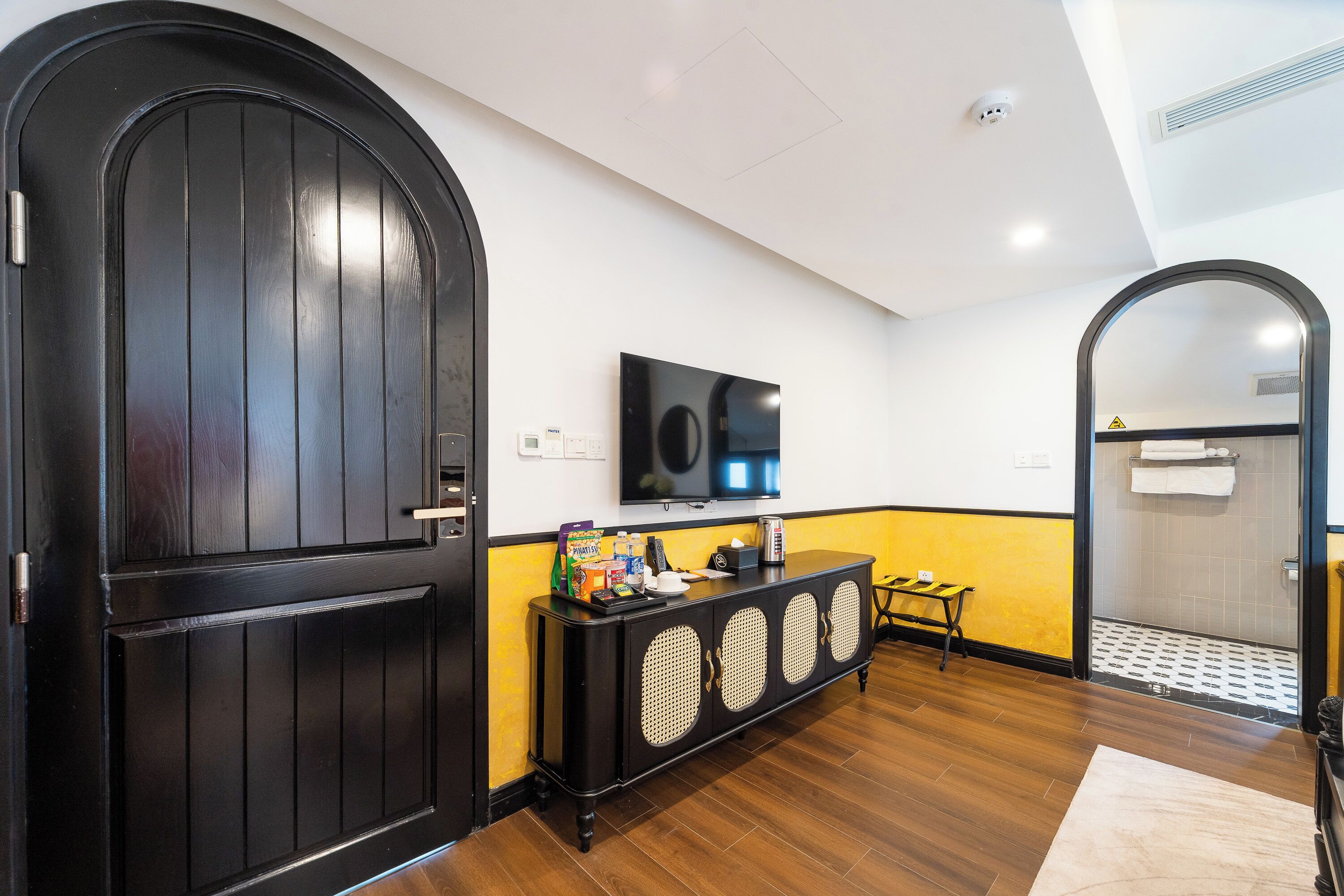Um saguão de hotel com uma grande porta preta, um aparador preto com diversos objetos, uma TV de tela plana na parede e uma parede amarela em destaque.