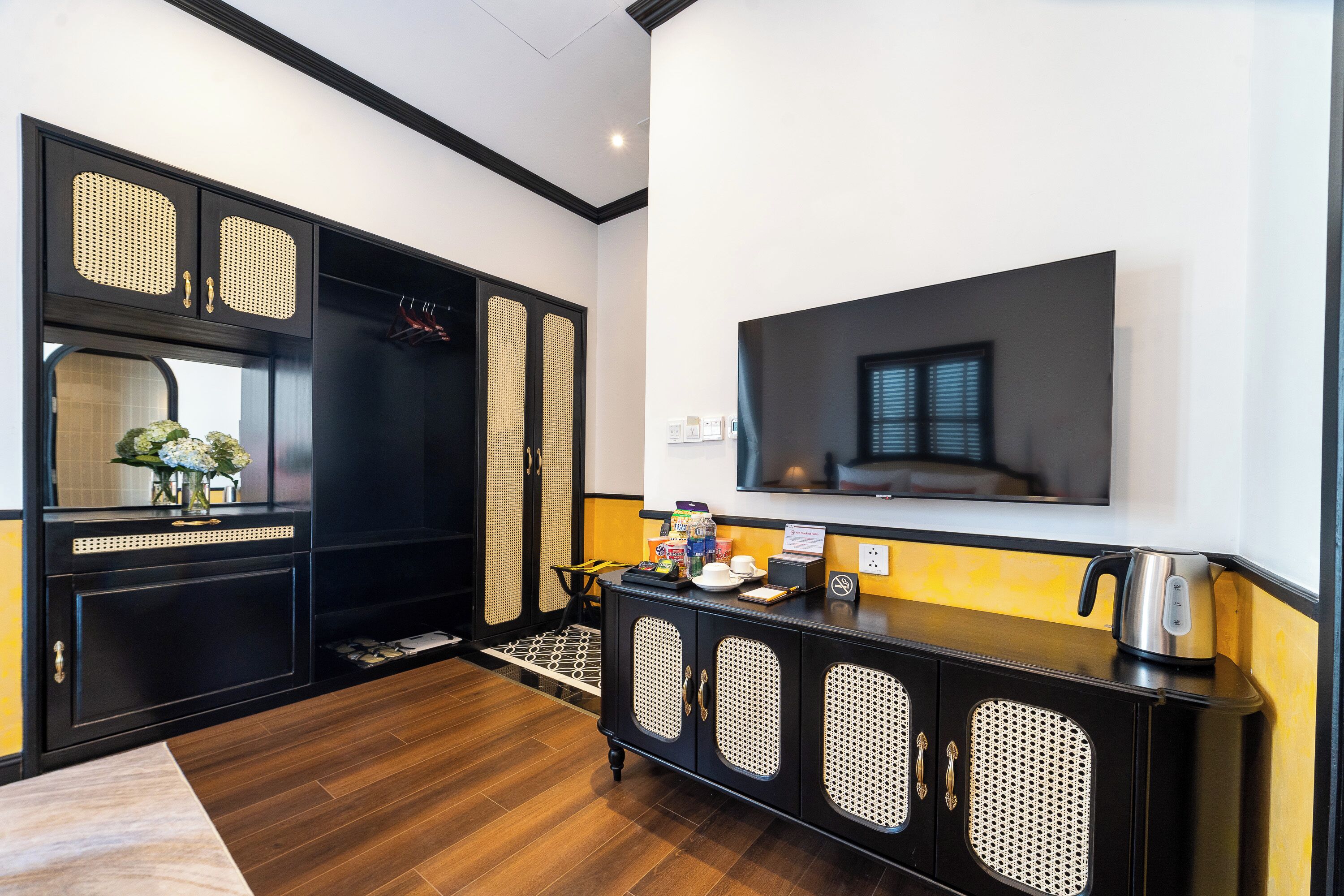 Quarto de hotel moderno com piso de madeira, esquema de cores preto e branco, TV de tela plana na parede e mesa de console preta com chaleira e outros objetos.