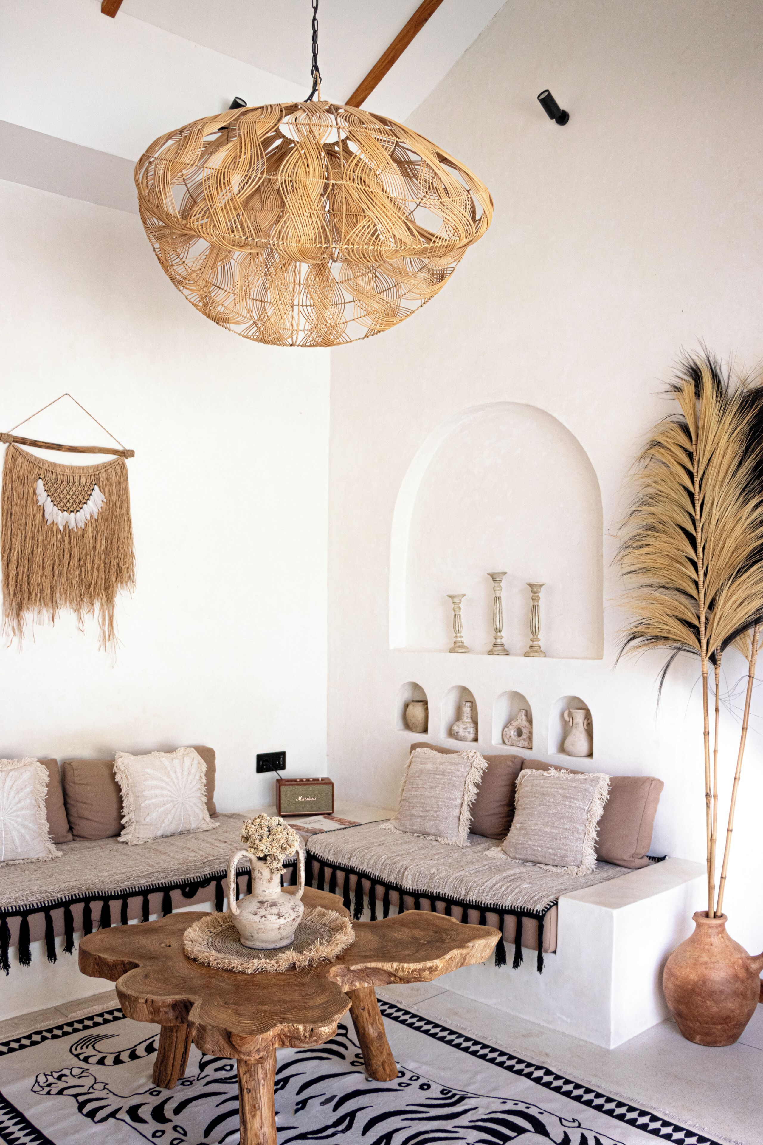 Uma sala de estar acolhedora com um candelabro de vime, um painel de parede em macramé, um sofá com almofadas, uma mesa de centro e um vaso com plantas secas.