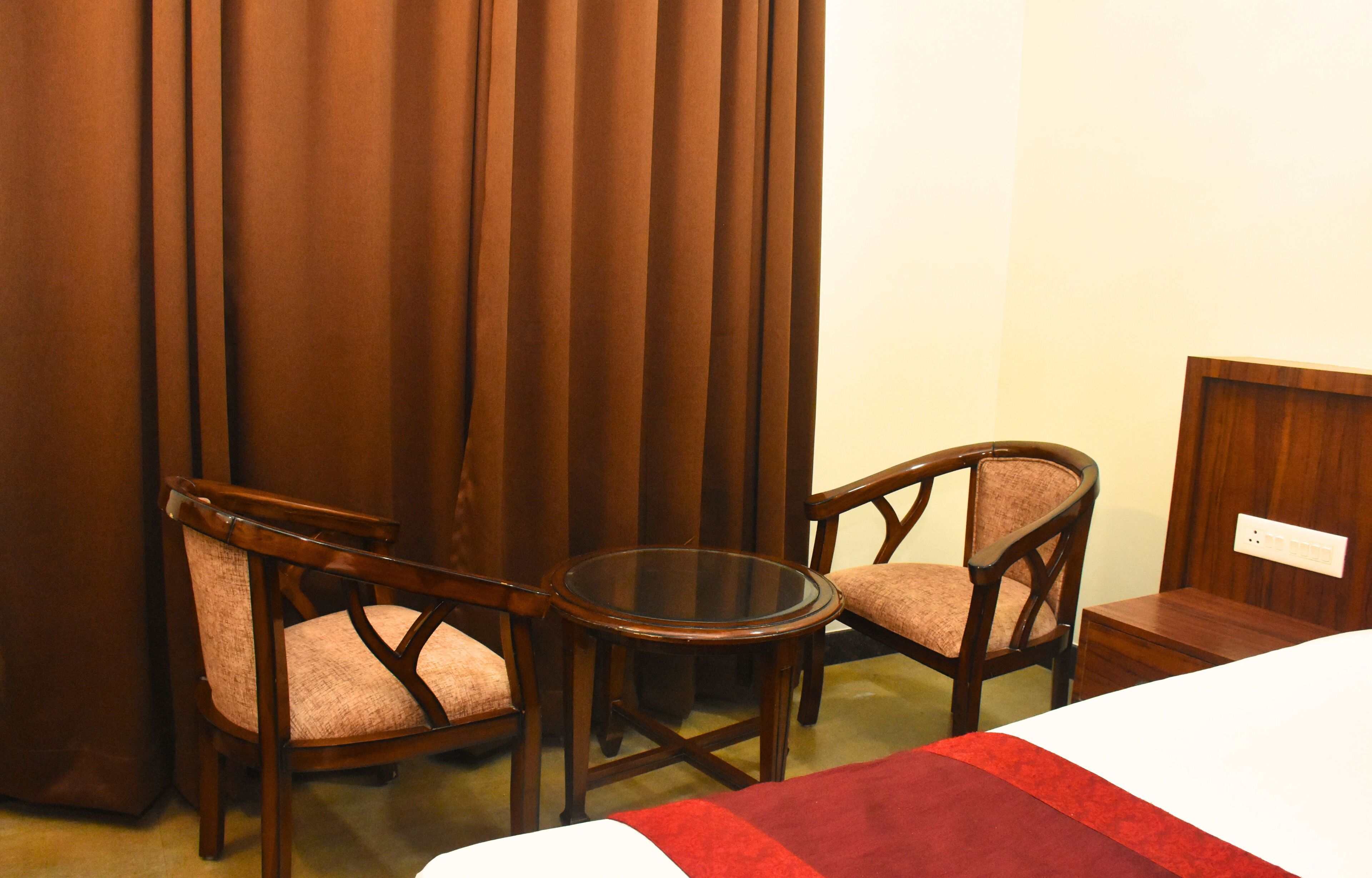 Quarto de hotel com cama, mesa redonda pequena, duas cadeiras e cortina.
