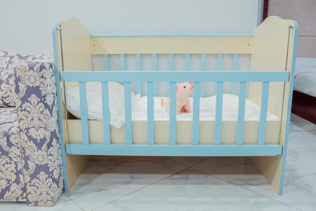 Um berço infantil com estrutura azul claro e colchão branco, posicionado sobre um chão de azulejos ao lado de um sofá estampado.