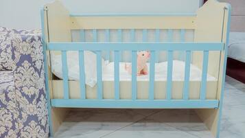 Um berço infantil com estrutura azul claro e colchão branco, posicionado sobre um chão de azulejos ao lado de um sofá estampado.