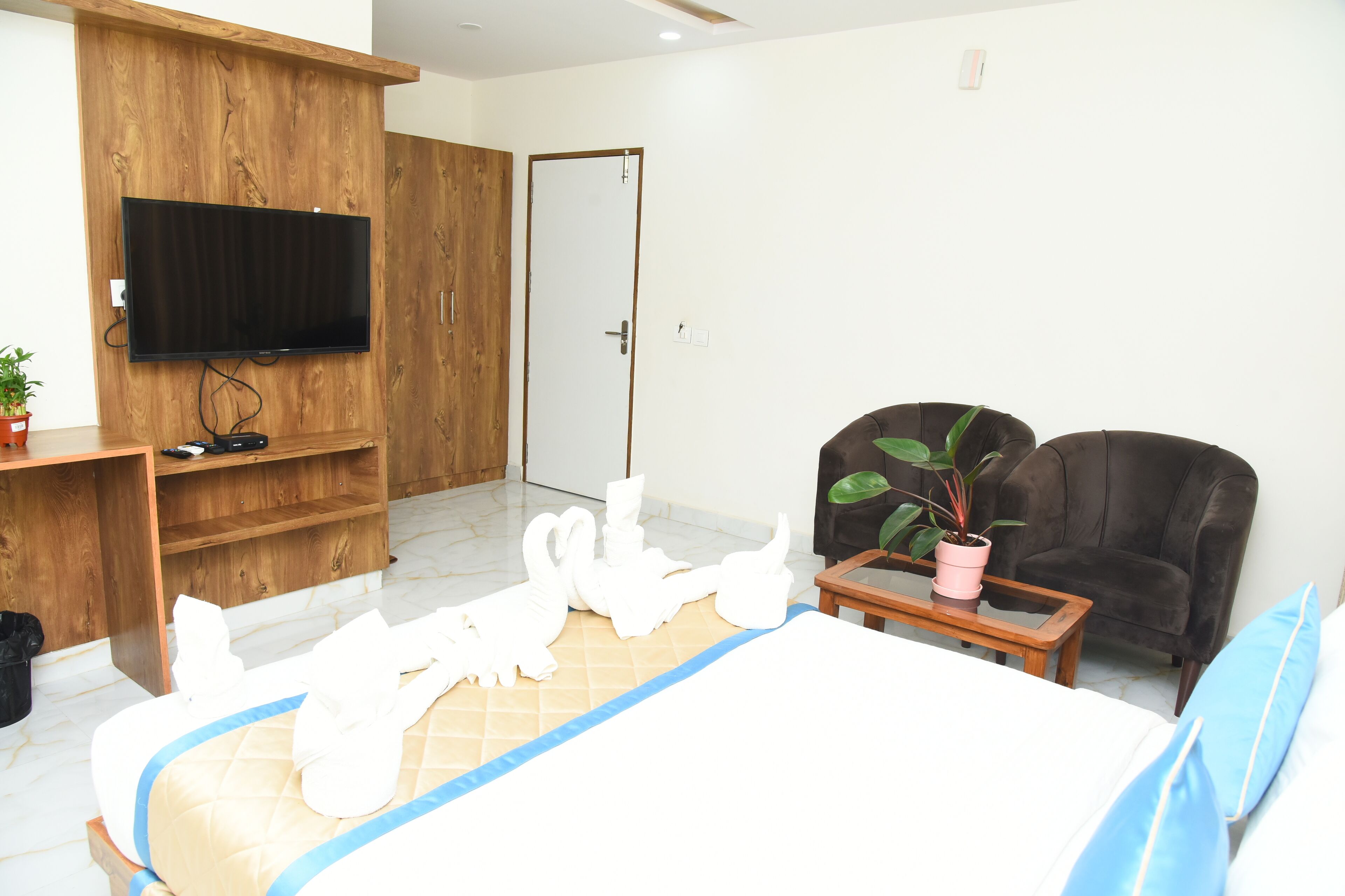 Um quarto com cama, TV fixada em um painel de madeira, poltrona escura, mesinha com planta e porta ao fundo.
