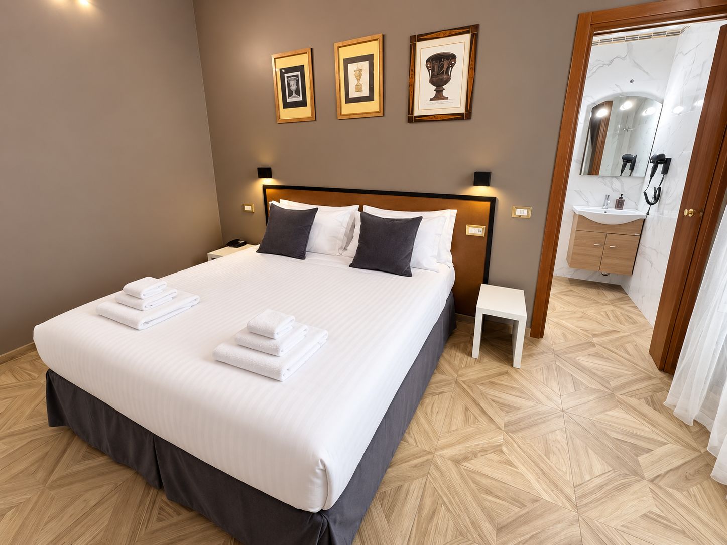 Quarto de hotel com cama grande, mesas de cabeceira, banheiro com espelho e pia, e quadros nas paredes.