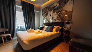 Quarto de hotel moderno com uma cama grande, piso de madeira, cabeceira com parede texturizada e uma cadeira perto da janela.
