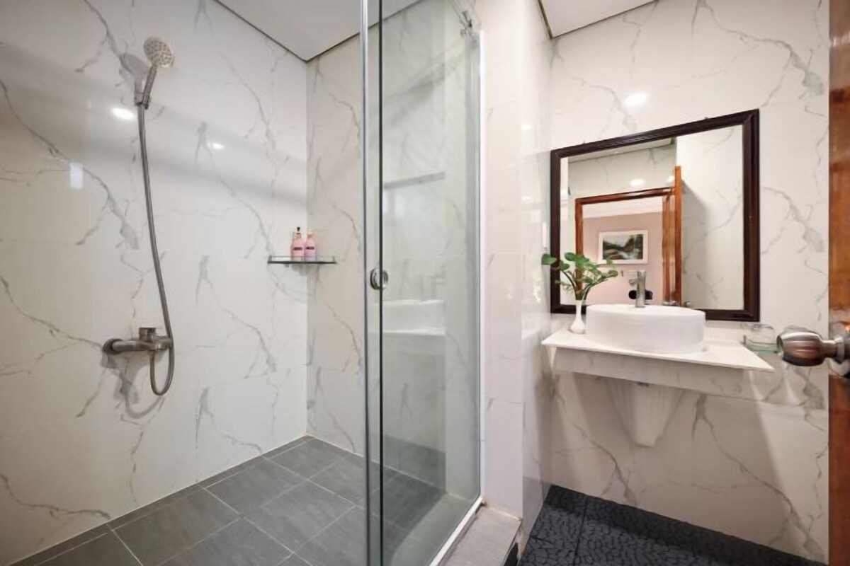 Casa de banho moderna com chuveiro envidraçado, parede revestida a mármore, lavatório branco com espelho e pequena prateleira com artigos de higiene pessoal.