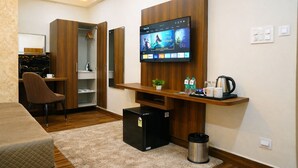 Quarto de hotel com televisão, mobiliário de madeira, uma secretária, uma cadeira, máquina de café e lixeira.