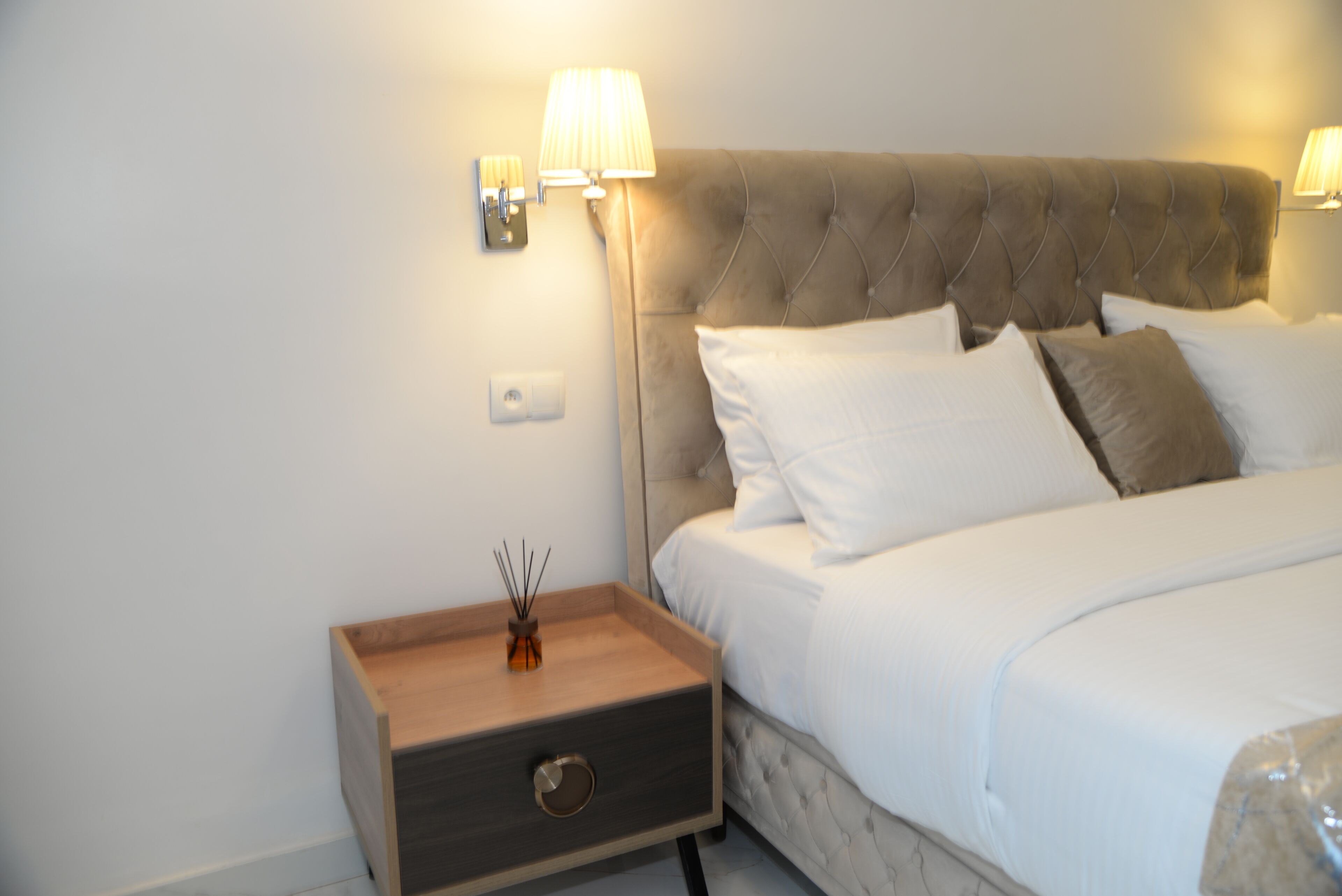 Cama bem arrumada com roupa de cama branca e castanha, mesa de cabeceira de madeira com gaveta e duas luminárias fixadas na parede.
