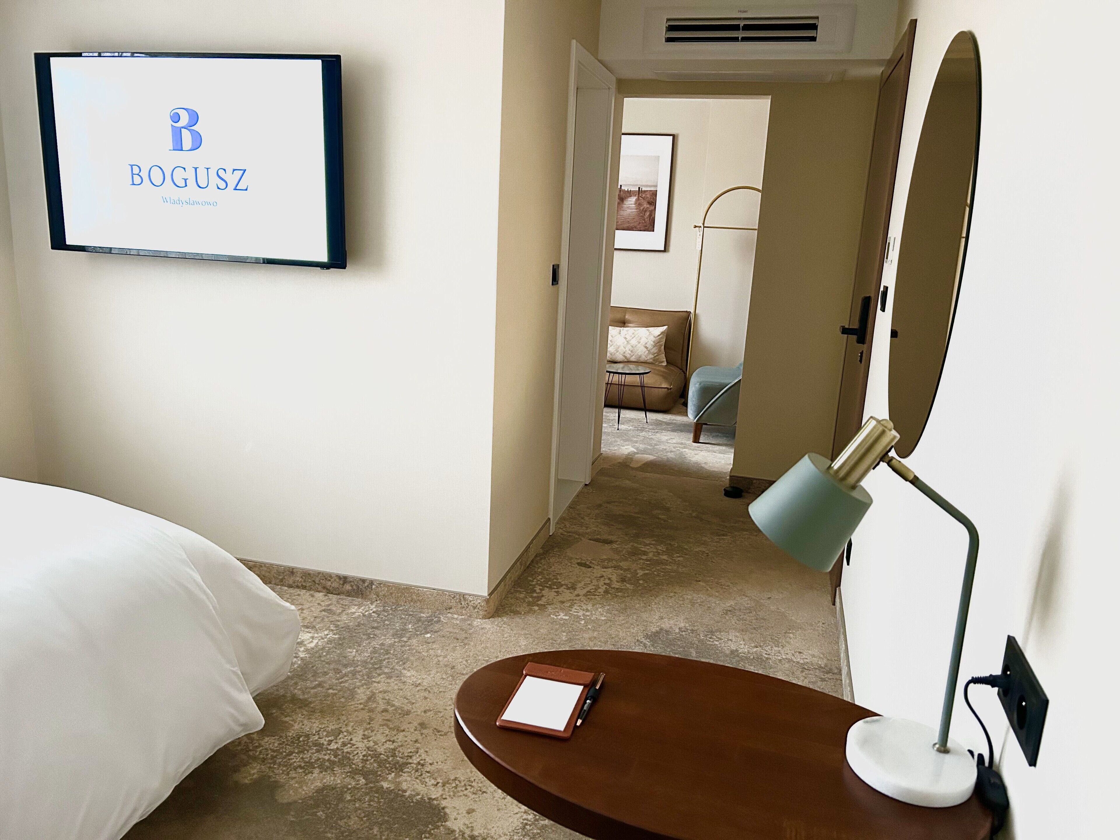 Quarto de hotel com uma cama, uma escrivaninha com abajur, um quadro na parede e um corredor que dá para outro quarto.