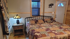 Um quarto aconchegante com uma cama de madeira, uma mesa de cabeceira com abajur, colcha acolchoada, janela com persianas e porta com adesivo de vaca.