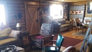 Interior acolhedor de uma cabana, com teto de madeira, um sofá, uma poltrona estampada, uma poltrona de couro e um espaço de jantar com mesa e cadeiras.