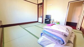 Um quarto japonês tradicional com tatames, uma cama com almofadas, uma televisão e uma pequena mesa com uma cadeira.