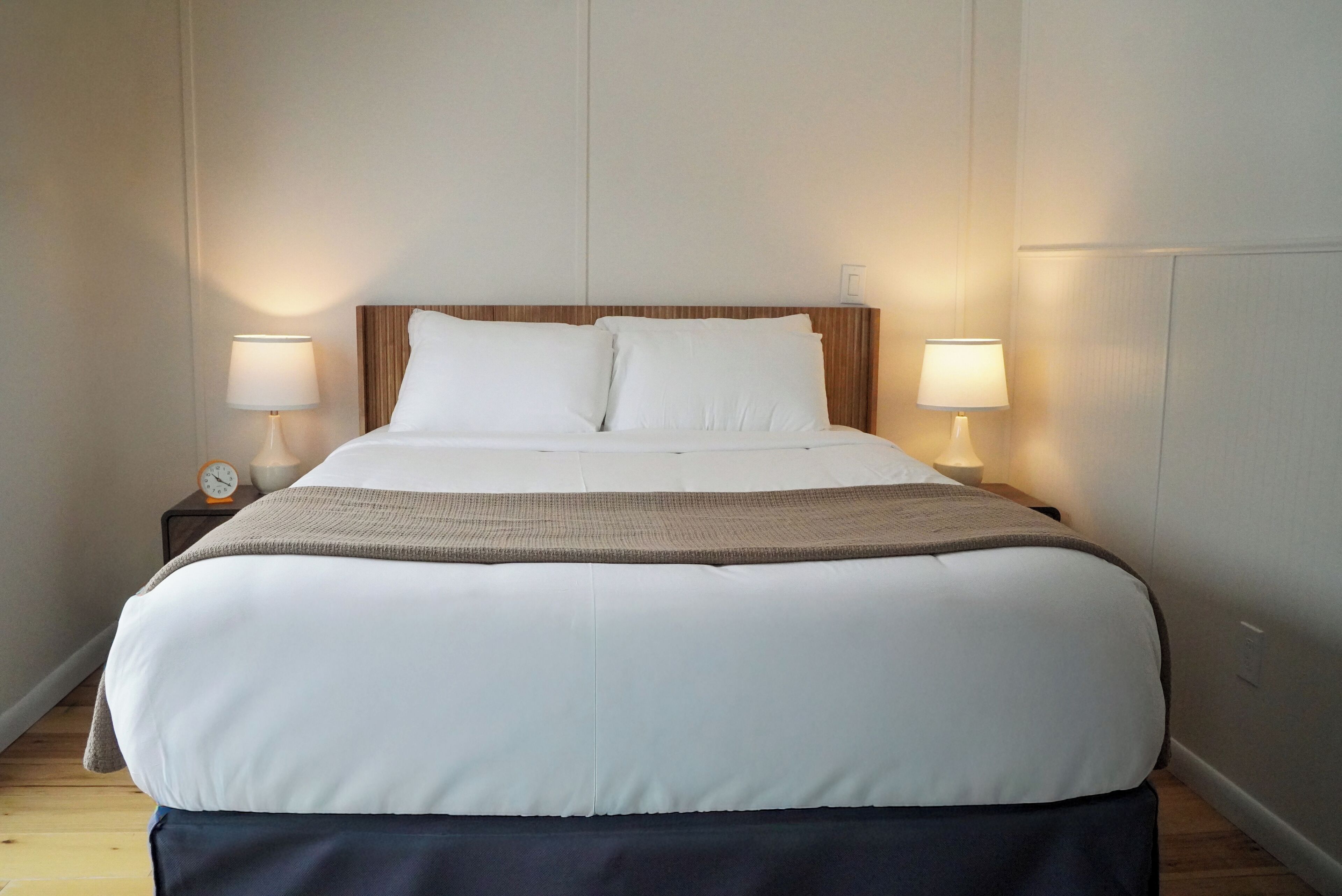 Uma cama bem arrumada com roupa de cama branca e manta marrom, com duas luminárias laterais em mesas de cabeceira combinando.