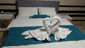 Uma cama bem arrumada com colcha azul e branca, duas toalhas brancas dispostas em formato de coração e um travesseiro azul na cabeceira.