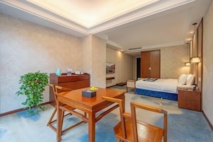 Quarto de hotel com mesa de jantar de madeira, cadeiras, cama, mesa de cabeceira, abajur e uma planta.