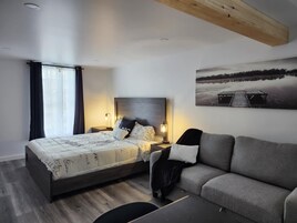 Quarto moderno com cama grande, sofá cinza, teto de madeira e pintura com vista para um lago.