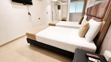 Quarto de hotel moderno com TV de tela plana na parede, cama com cabeceira, mesa de cabeceira com controle remoto e banheiro visível através de uma porta aberta.