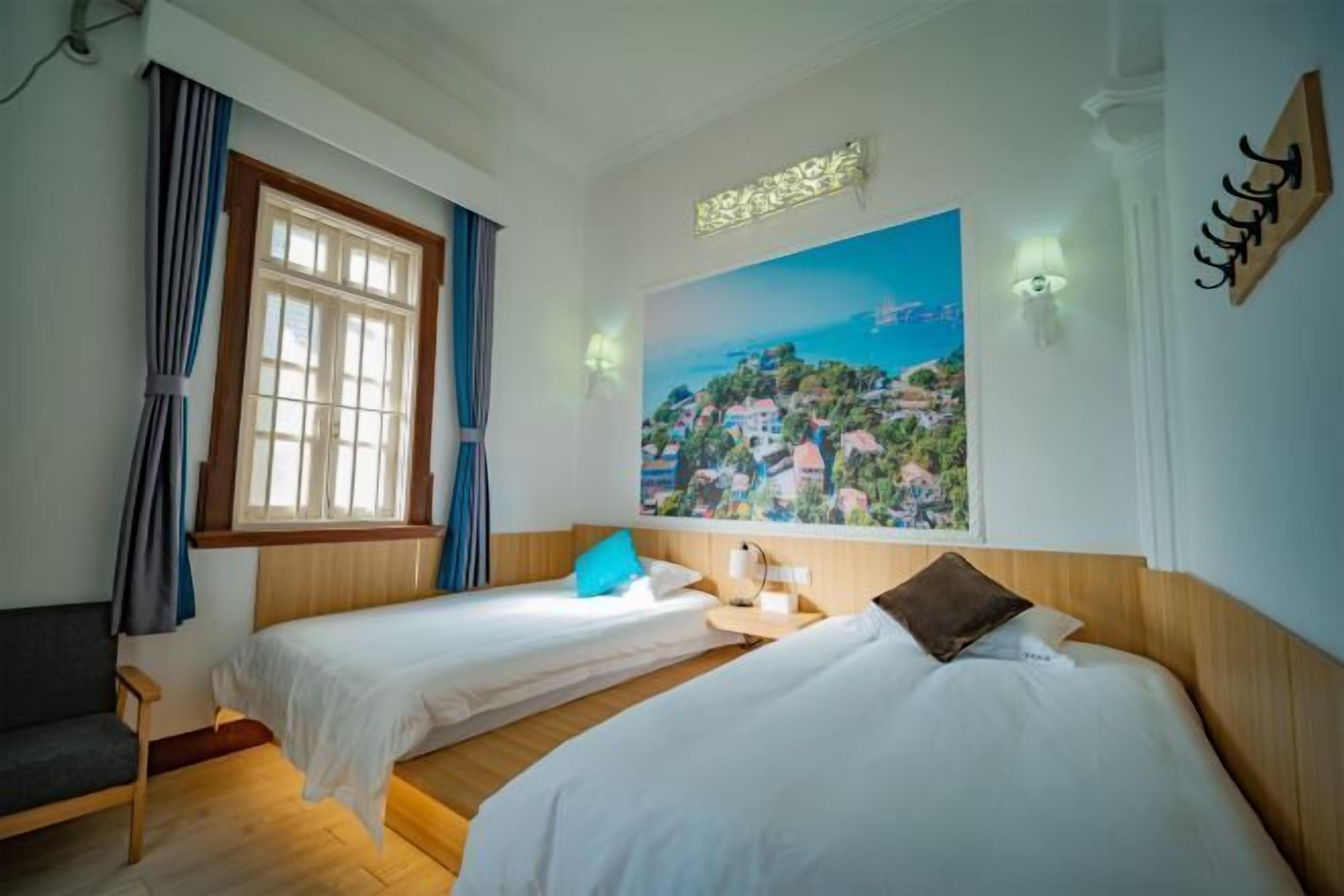 Um quarto com duas camas, uma janela com cortinas, um quadro de uma vila costeira e um abajur na parede.