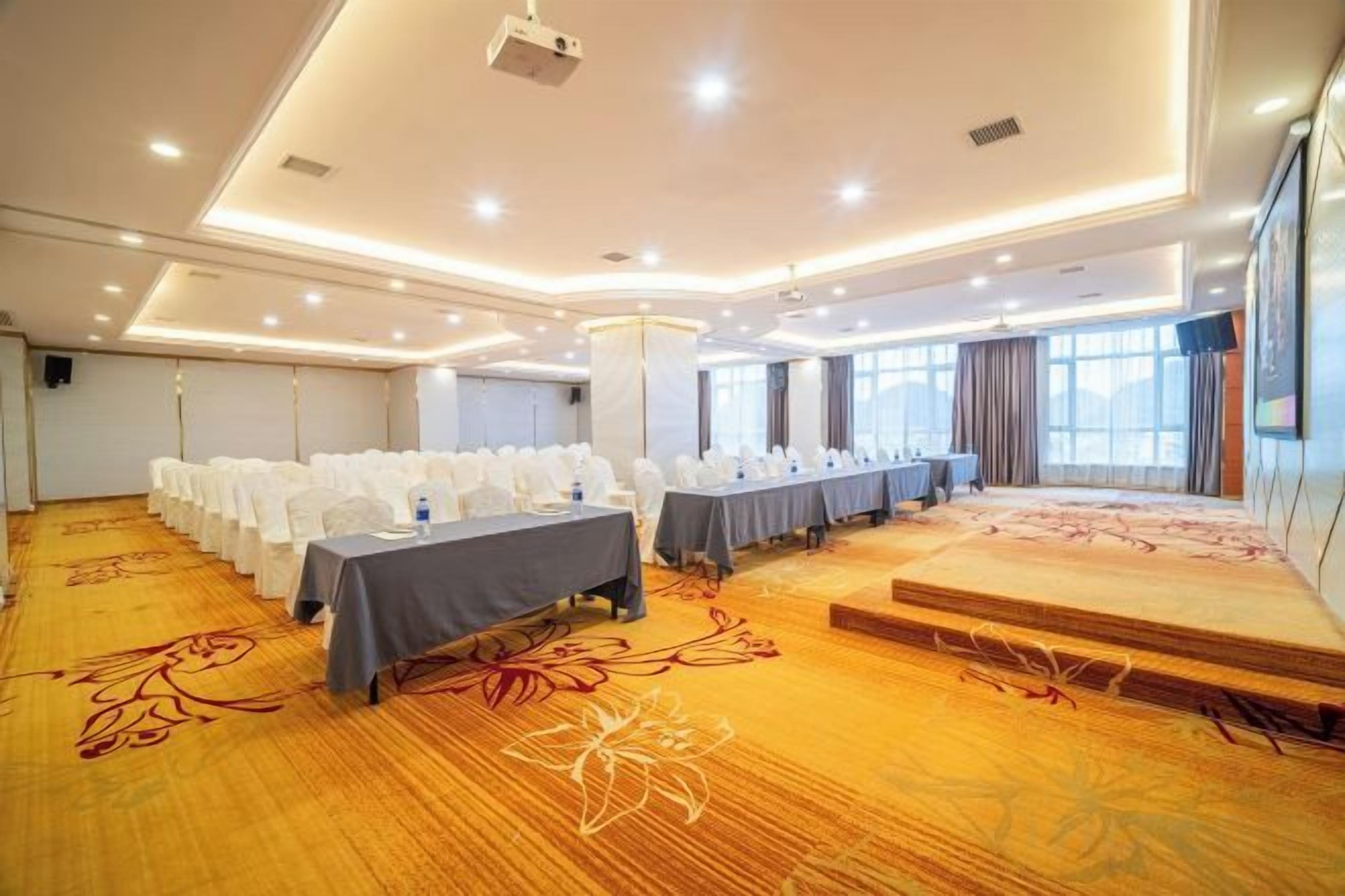 Uma sala de conferência com mesas dispostas em fileiras, palco e amplas janelas.