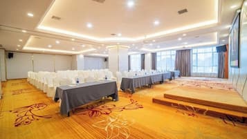 Uma sala de conferência com mesas dispostas em fileiras, palco e amplas janelas.