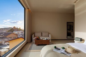 Quarto de hotel com uma janela grande, uma cama, um sofá, uma mesa de centro com frutas e uma mesinha com um livro e um chapéu.