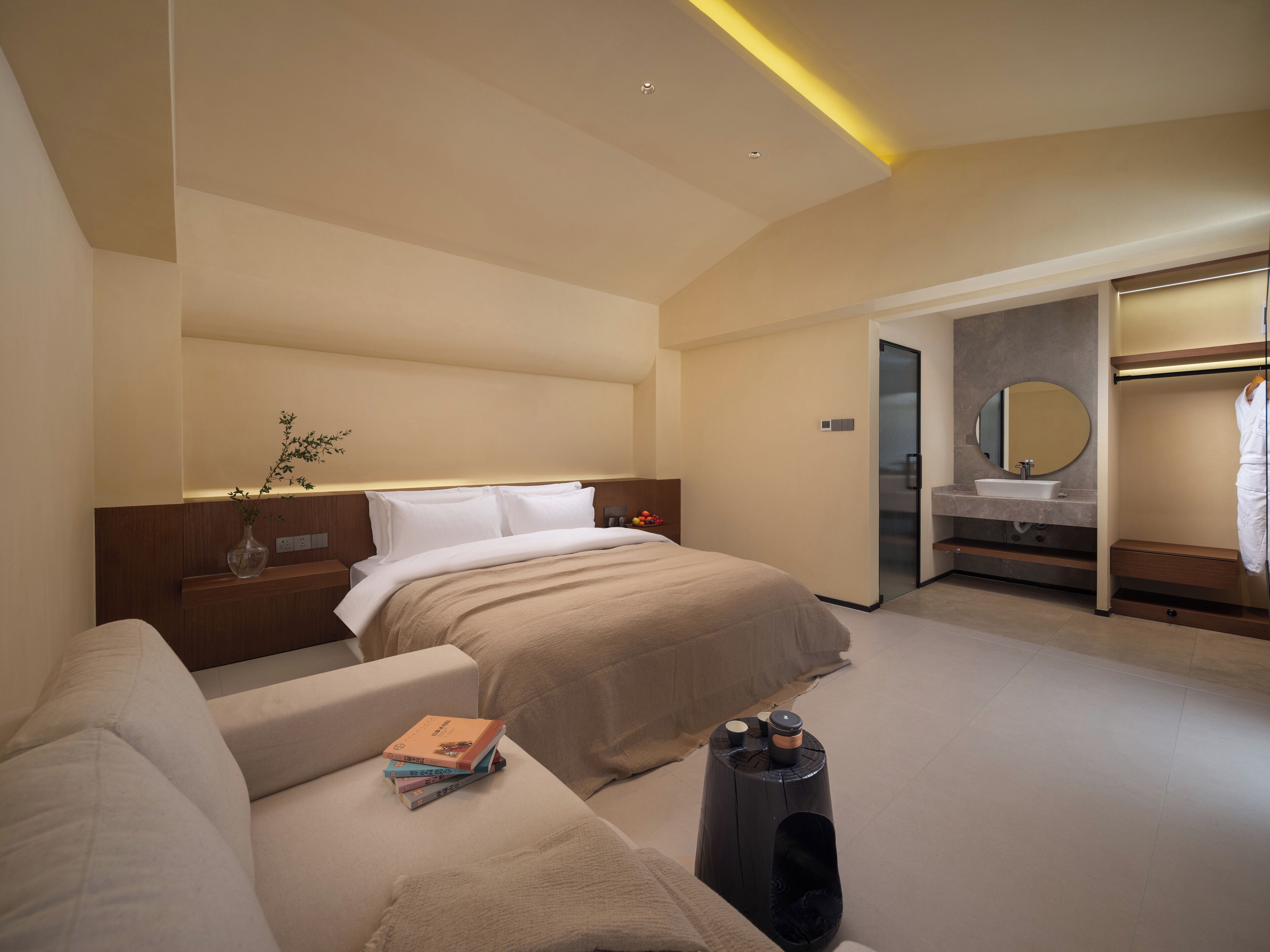 Quarto de hotel moderno com cama, sofá, mesa pequena com um vaso e espelho sobre a pia.