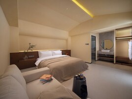 Quarto de hotel moderno com cama, sofá, mesa pequena com um vaso e espelho sobre a pia.