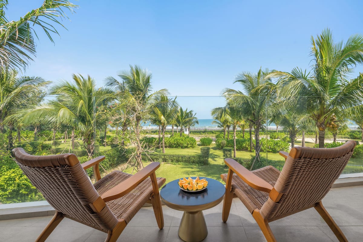 Varanda com cadeiras de vime e uma mesa, com vista para um jardim exuberante e o mar.