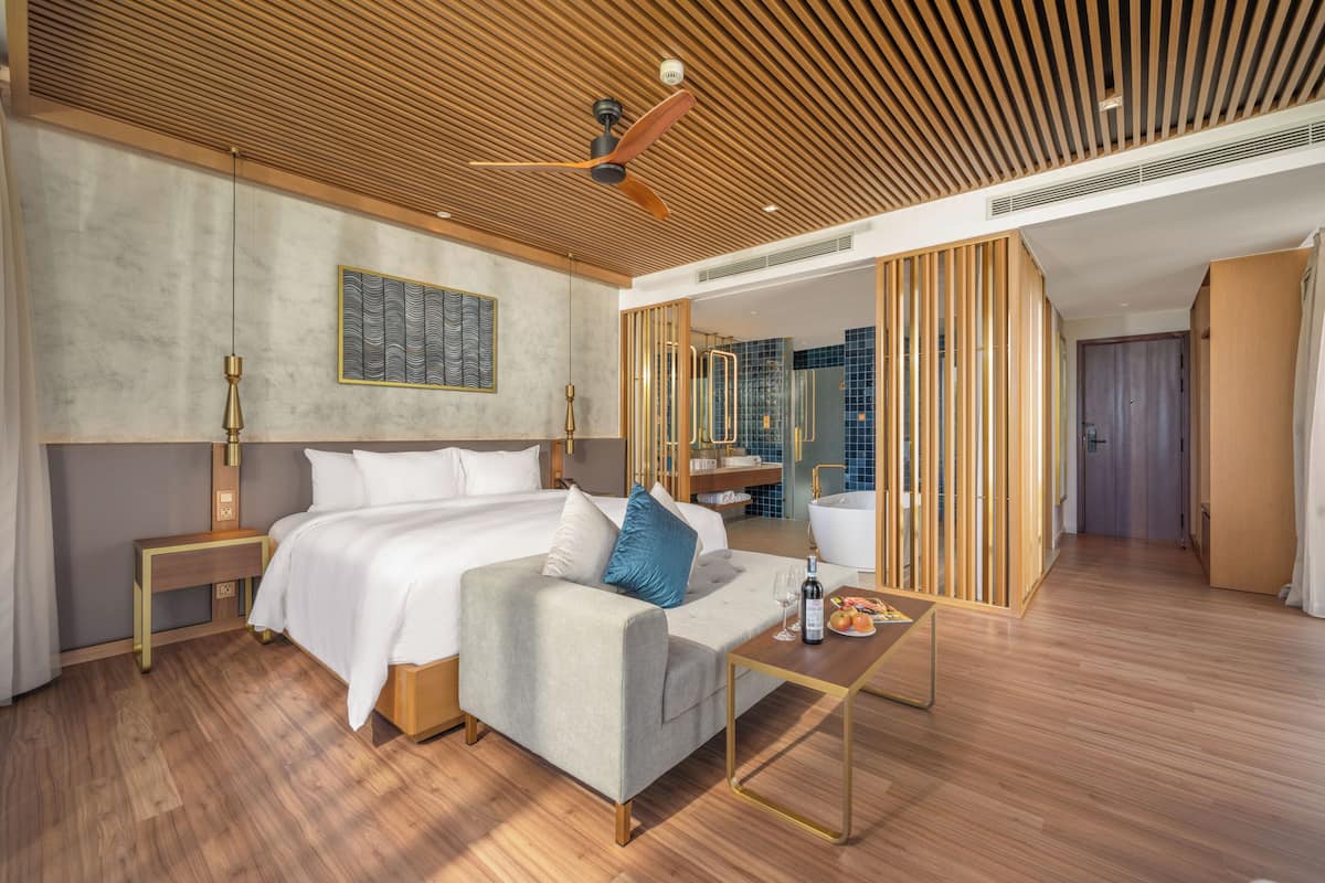 Quarto de hotel moderno com cama, sofá, mesa pequena com garrafa e copos, ventoinha no teto e banheiro visível por uma porta deslizante.
