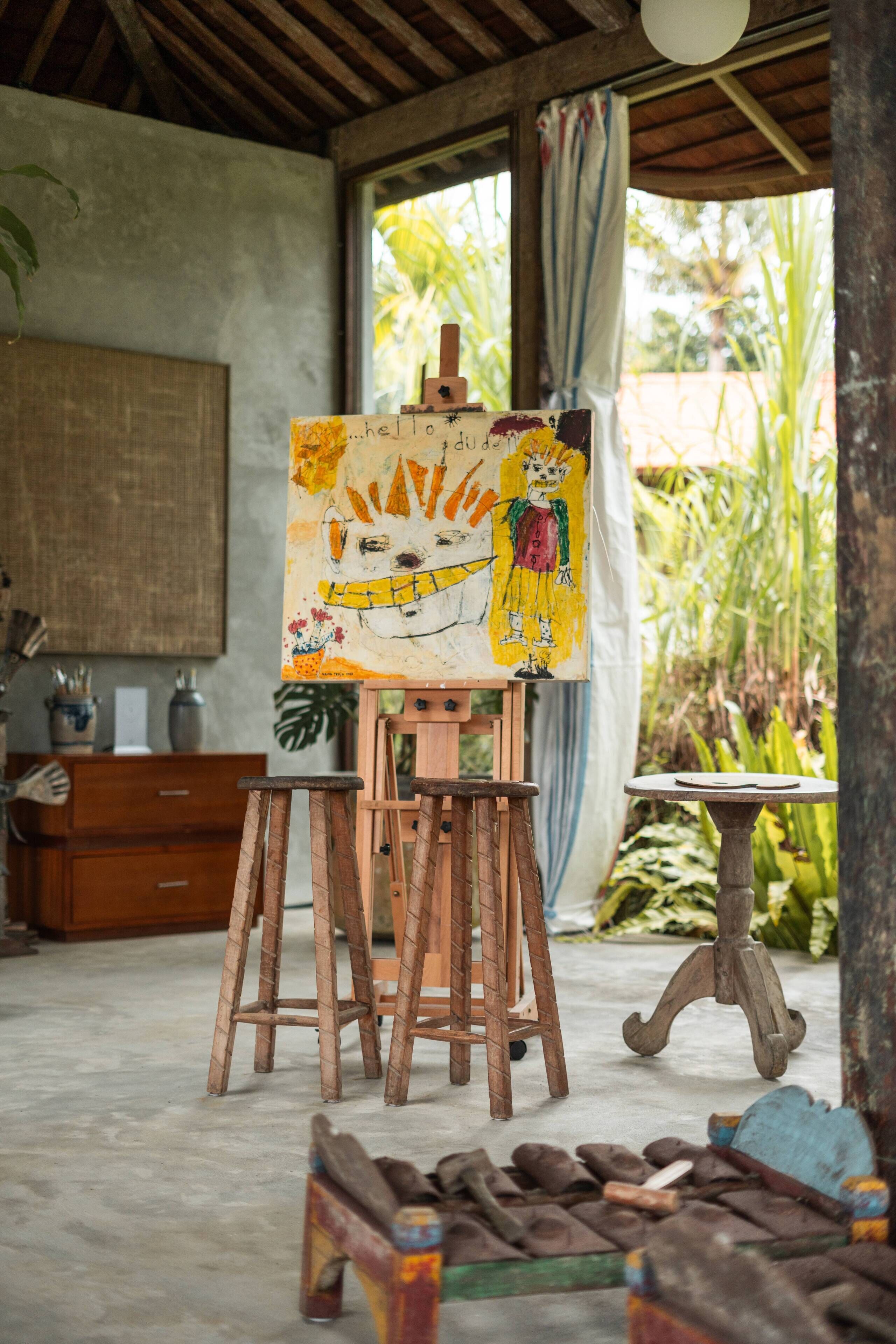 Um atelier de artista com um cavalete exibindo uma pintura colorida, bancos de madeira e uma pequena mesa.