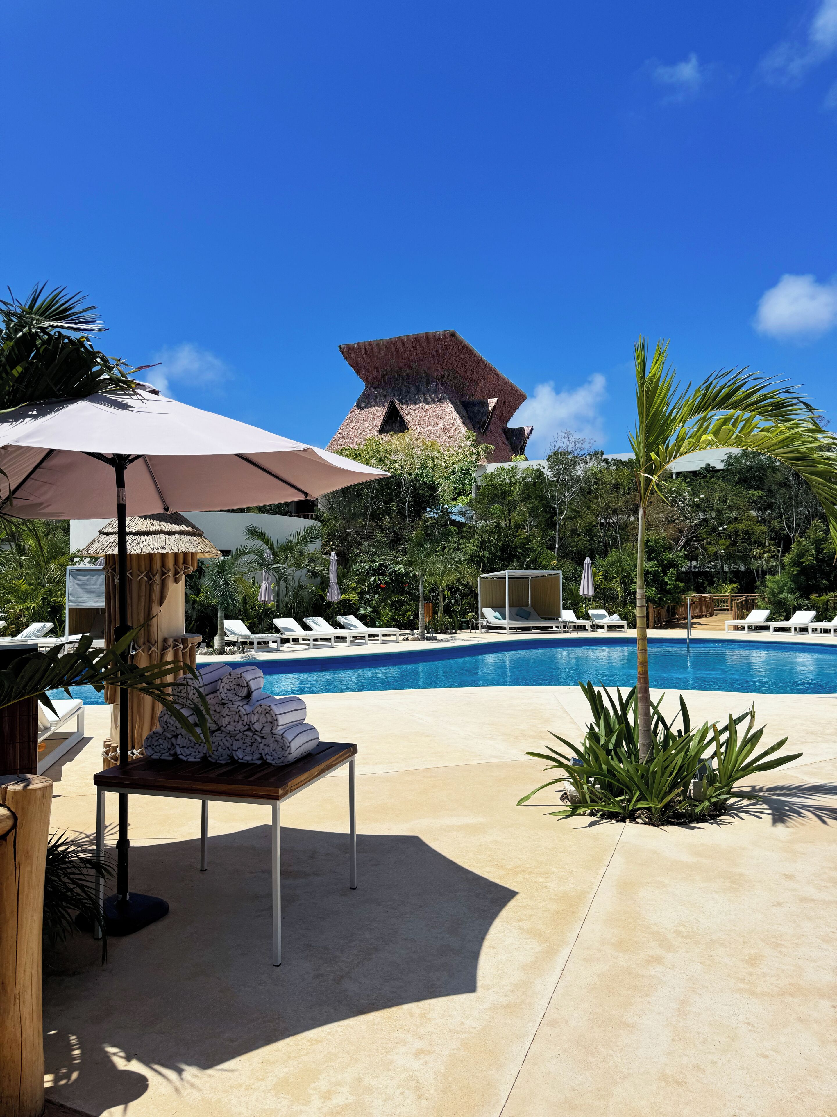 Uma área à beira da piscina com espreguiçadeiras, uma mesa com toalhas e uma estrutura com telhado de palha.