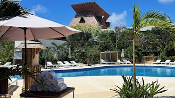 Uma área à beira da piscina com espreguiçadeiras, uma mesa com toalhas e uma estrutura com telhado de palha.