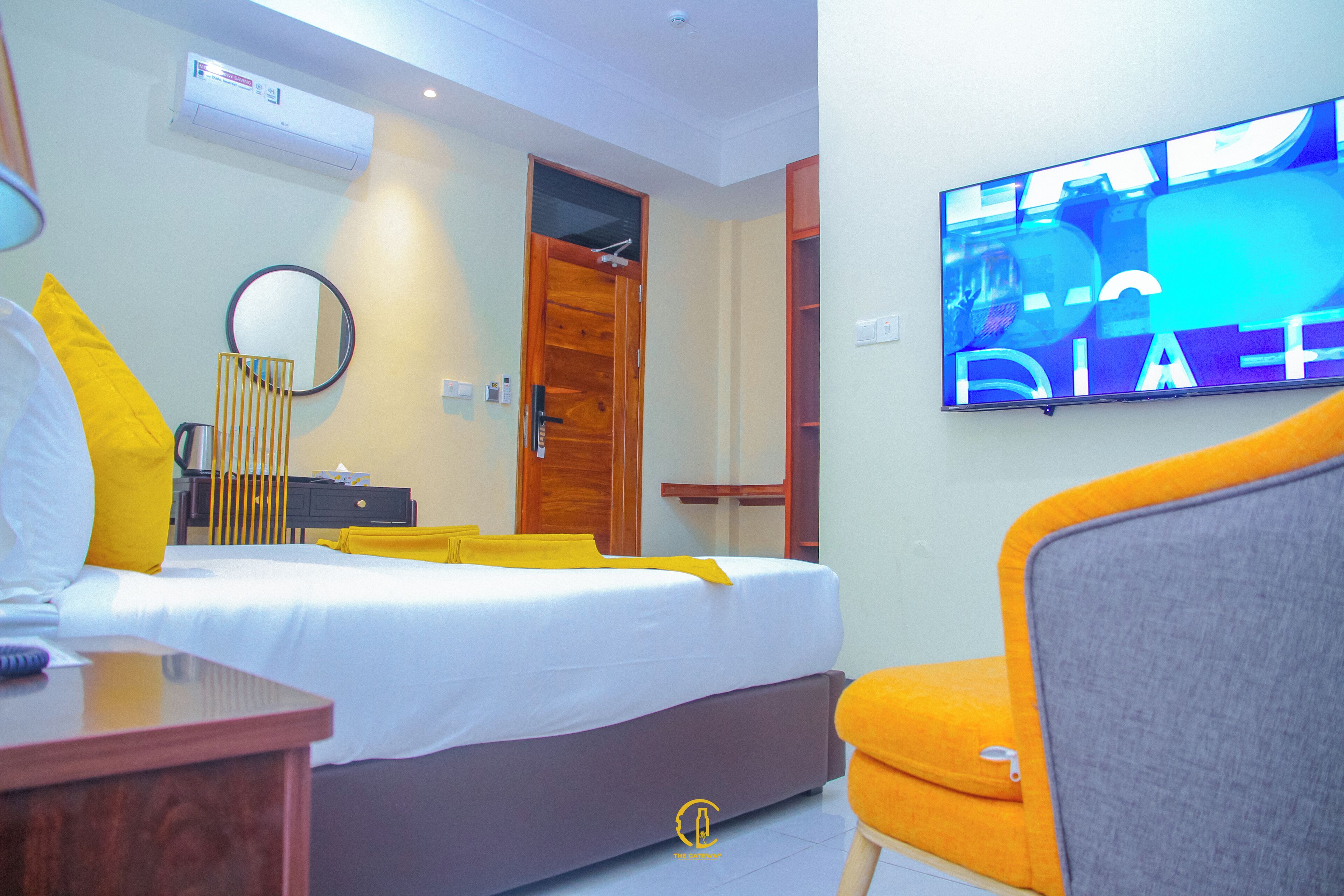 Quarto de hotel com cama, escrivaninha, cadeira, televisão e guarda-roupa.