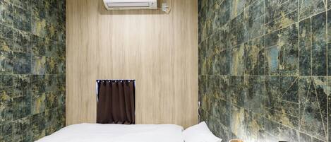 Quarto de hotel com cama, ar-condicionado fixado na parede, cortina, mesa de cabeceira com abajur e cafeteira.