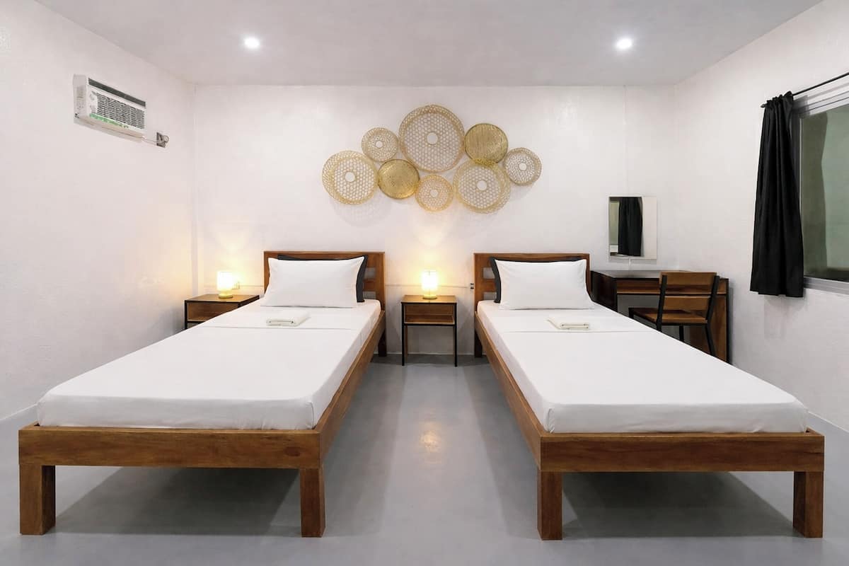 Quarto com duas camas de madeira, uma escrivaninha pequena, ar condicionado fixado na parede e um painel decorativo.