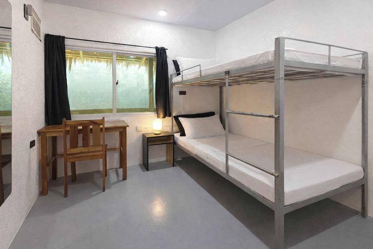 Quarto com beliche, escrivaninha, cadeira e janela com cortinas.