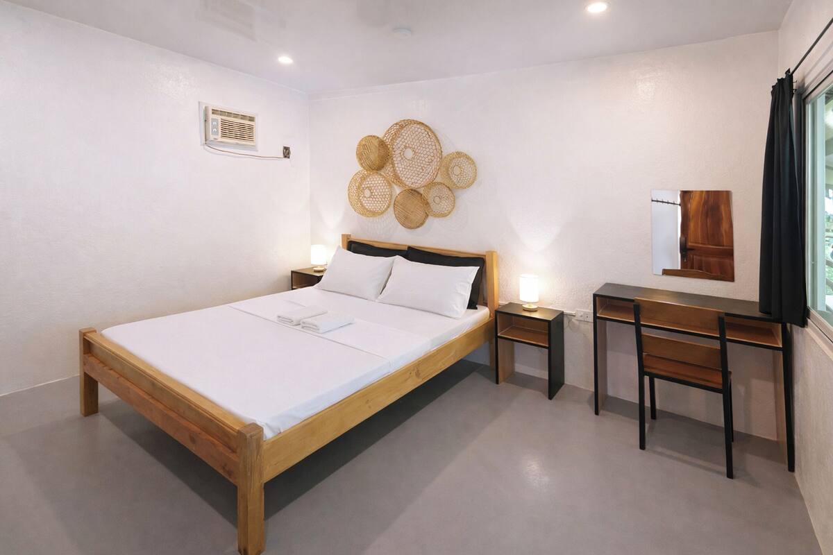 Quarto com cama de madeira, mesas de cabeceira, escrivaninha e um objeto decorativo fixado na parede.