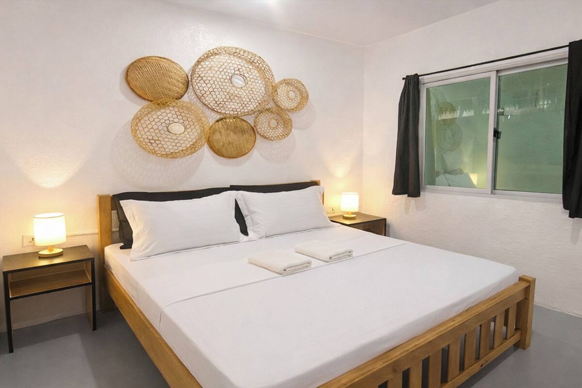 Cama bem arrumada com lençóis brancos, cabeceira e peseira de madeira, acompanhadas por duas mesas de cabeceira com abajures.