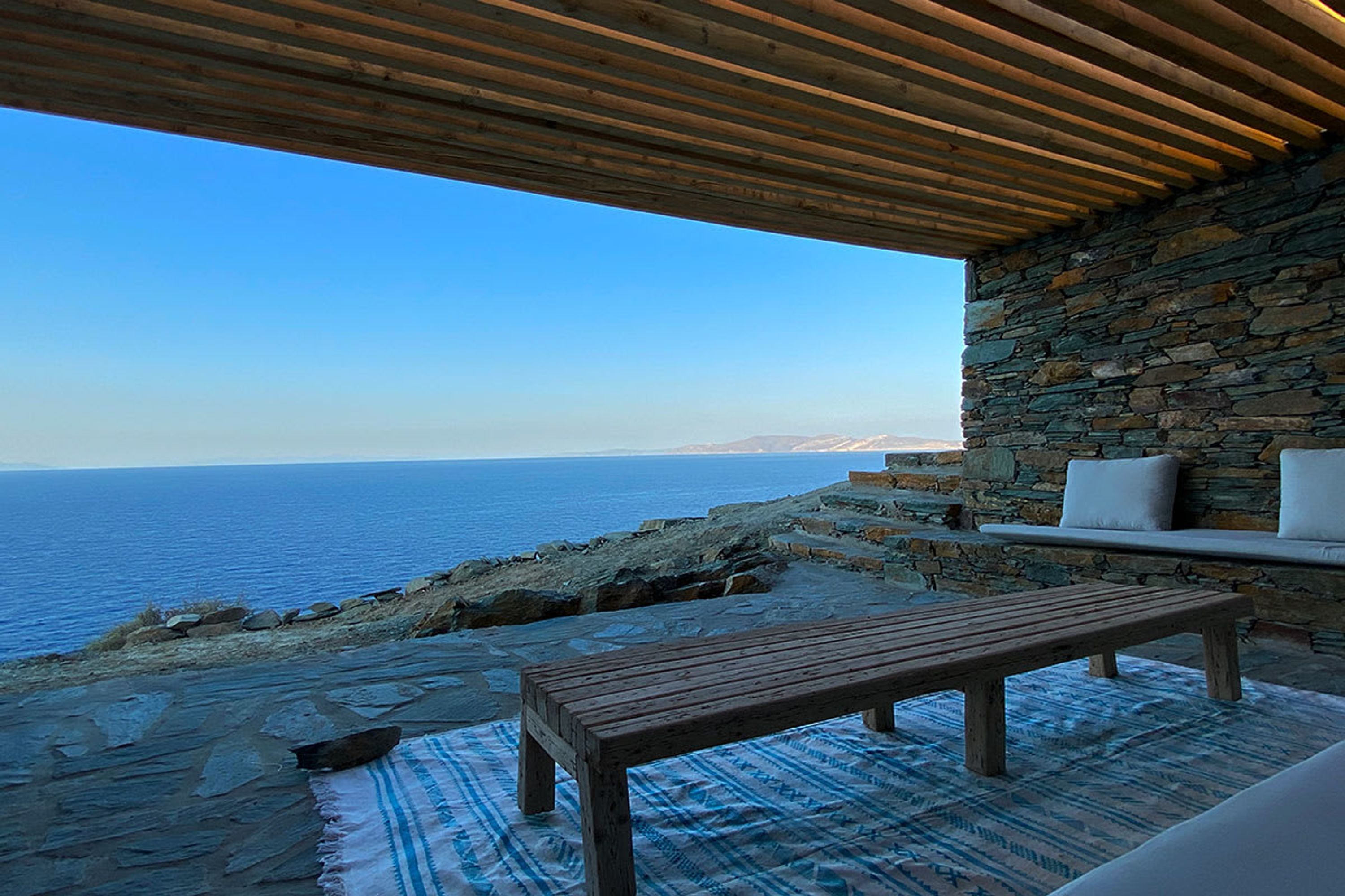 Quarto com paredes de pedra, teto de madeira, um banco e vista para o mar.