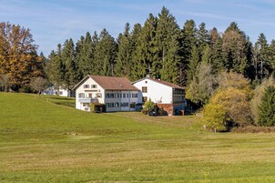 Um edifício grande e branco com várias janelas e telhado inclinado, rodeado por um campo gramado e floresta densa.