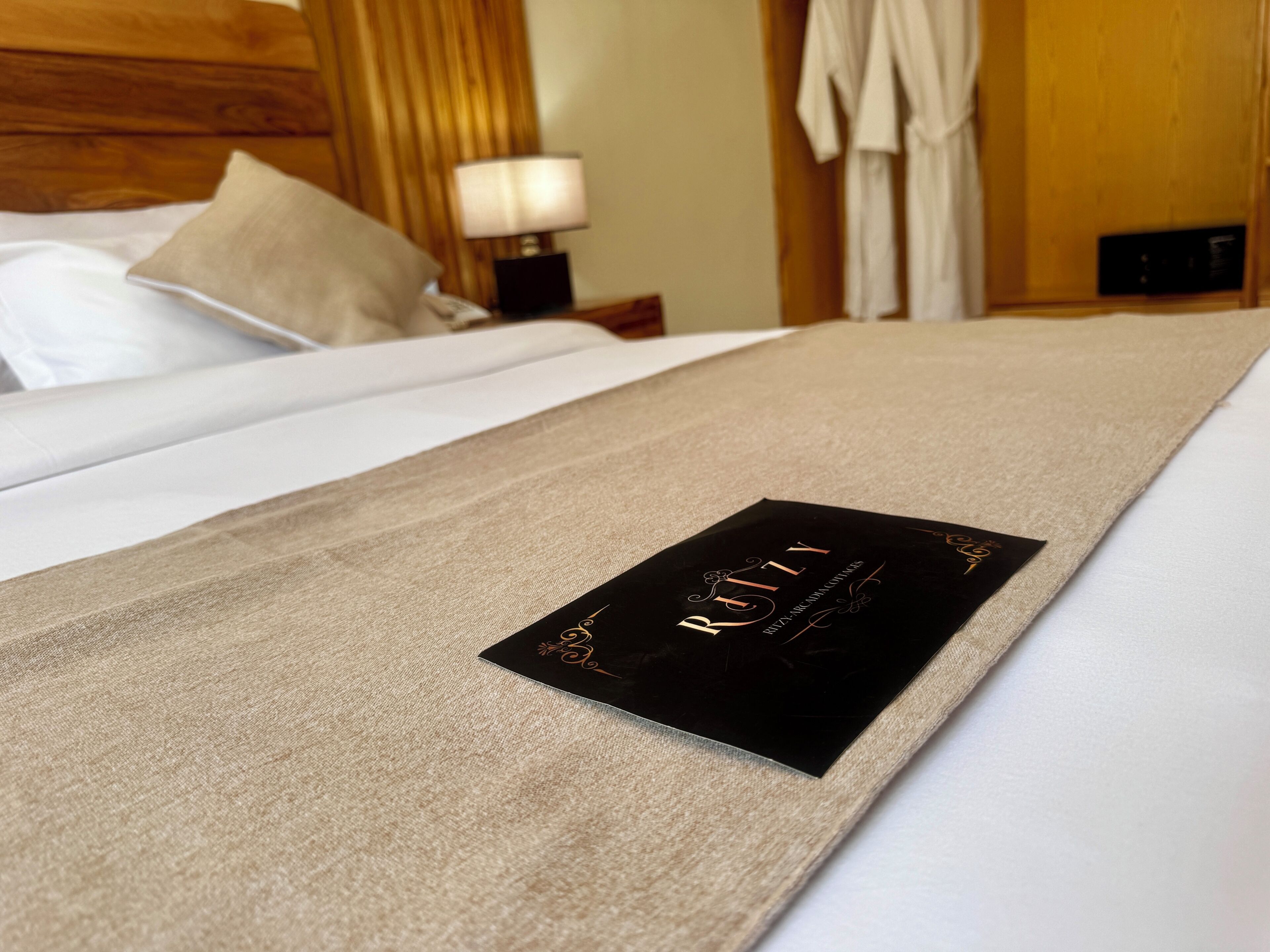Uma cama bem arrumada com uma placa preta sobre o lençol, cabeceira de madeira, luminária de cabeceira e cortina branca.