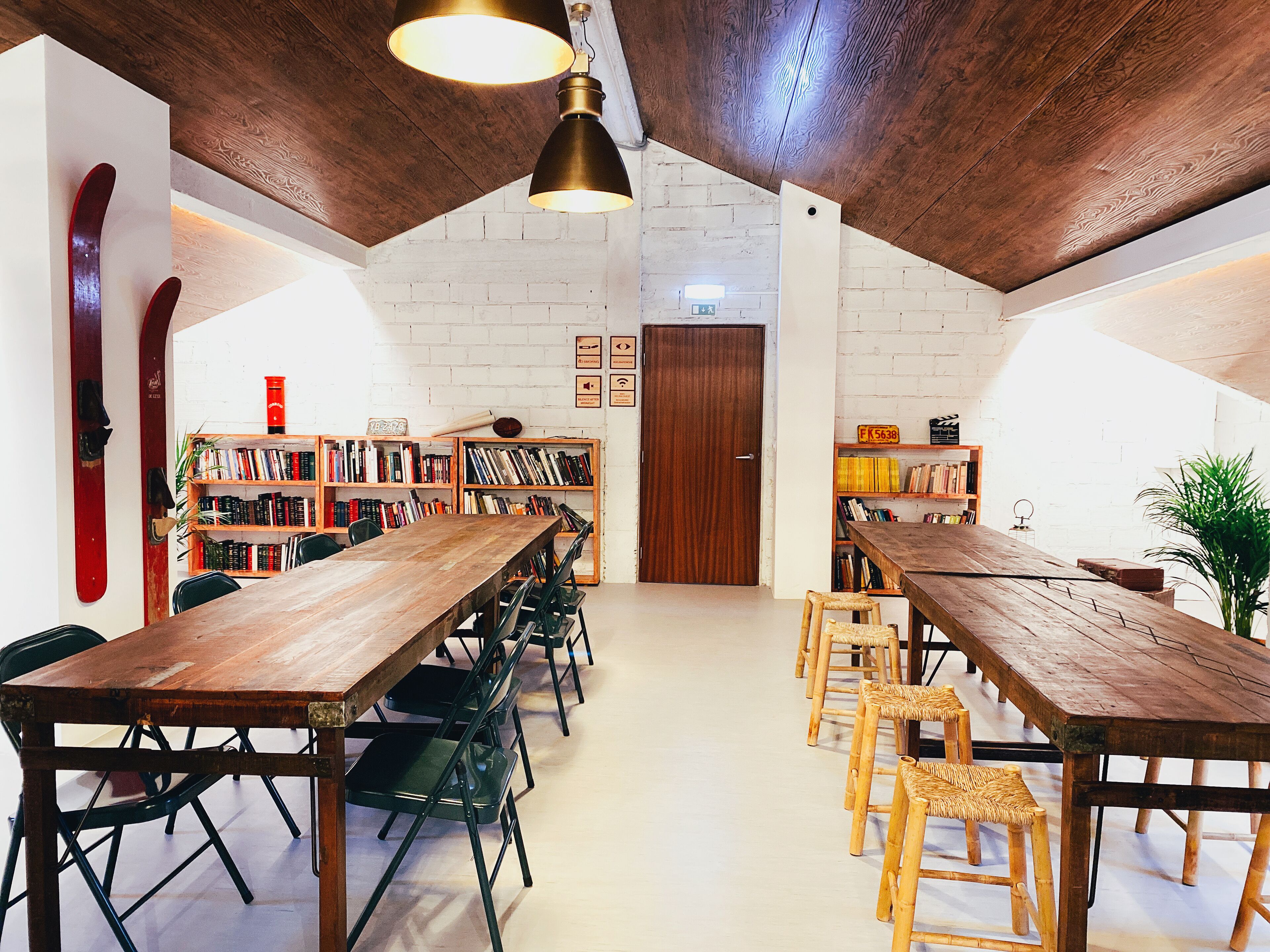 Um quarto espaçoso com tetos altos, vigas de madeira aparentes e uma longa mesa de madeira rodeada por cadeiras. Há várias estantes repletas de livros, e uma porta com uma placa indicando a saída.