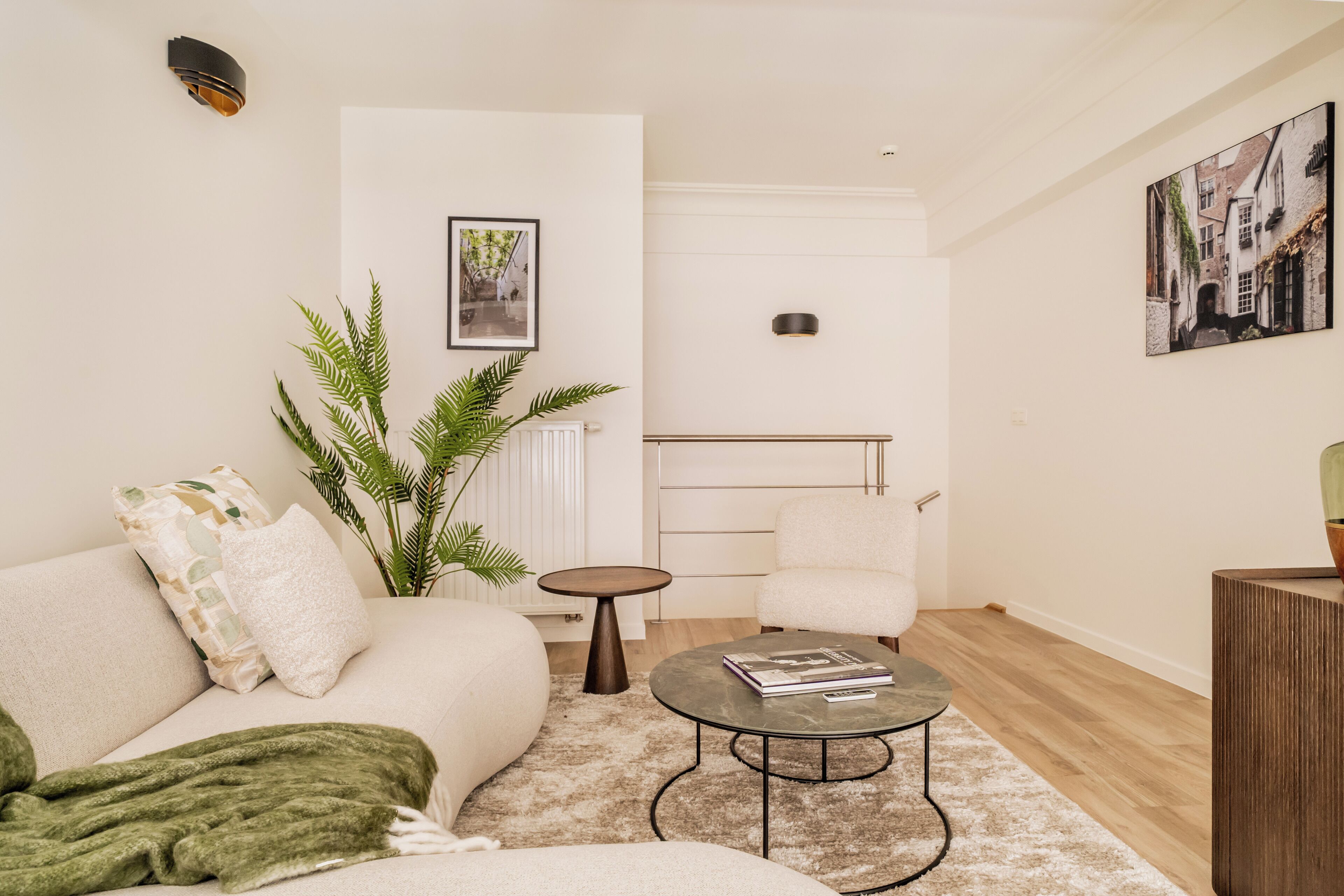 Uma sala de estar moderna com um sofá, uma mesa de centro, uma mesinha lateral e uma poltrona. Há uma planta, um quadro na parede e um radiador.