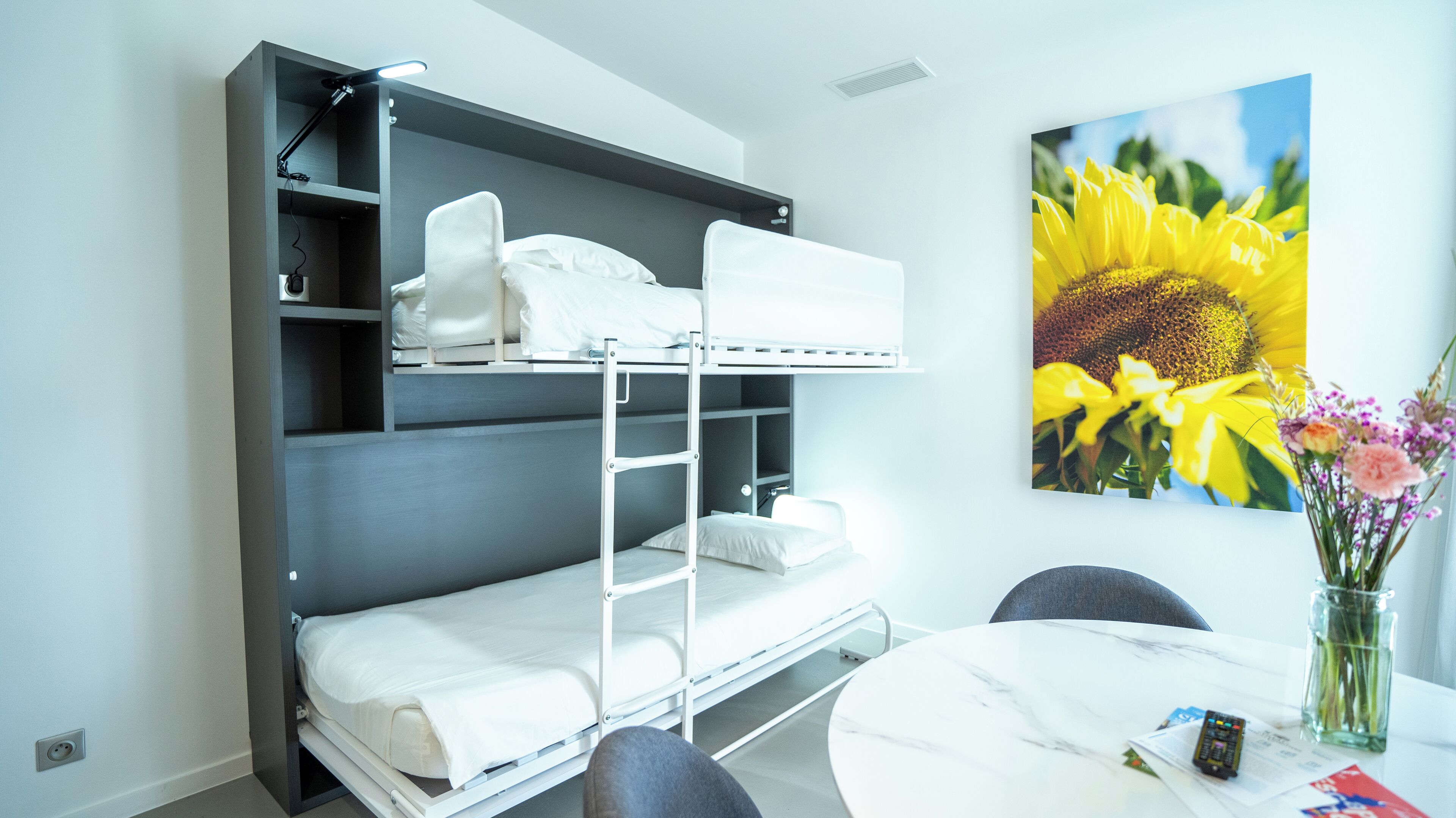 Quarto com beliche, uma escrivaninha e cadeira, um vaso com flores e um quadro de girassol na parede.