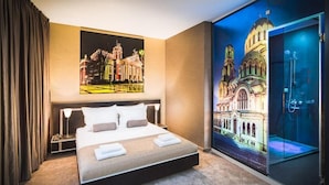 Quarto de hotel com uma cama grande, uma mesa de cabeceira, uma televisão na parede exibindo uma vista urbana e uma porta de vidro que dá para o banheiro.