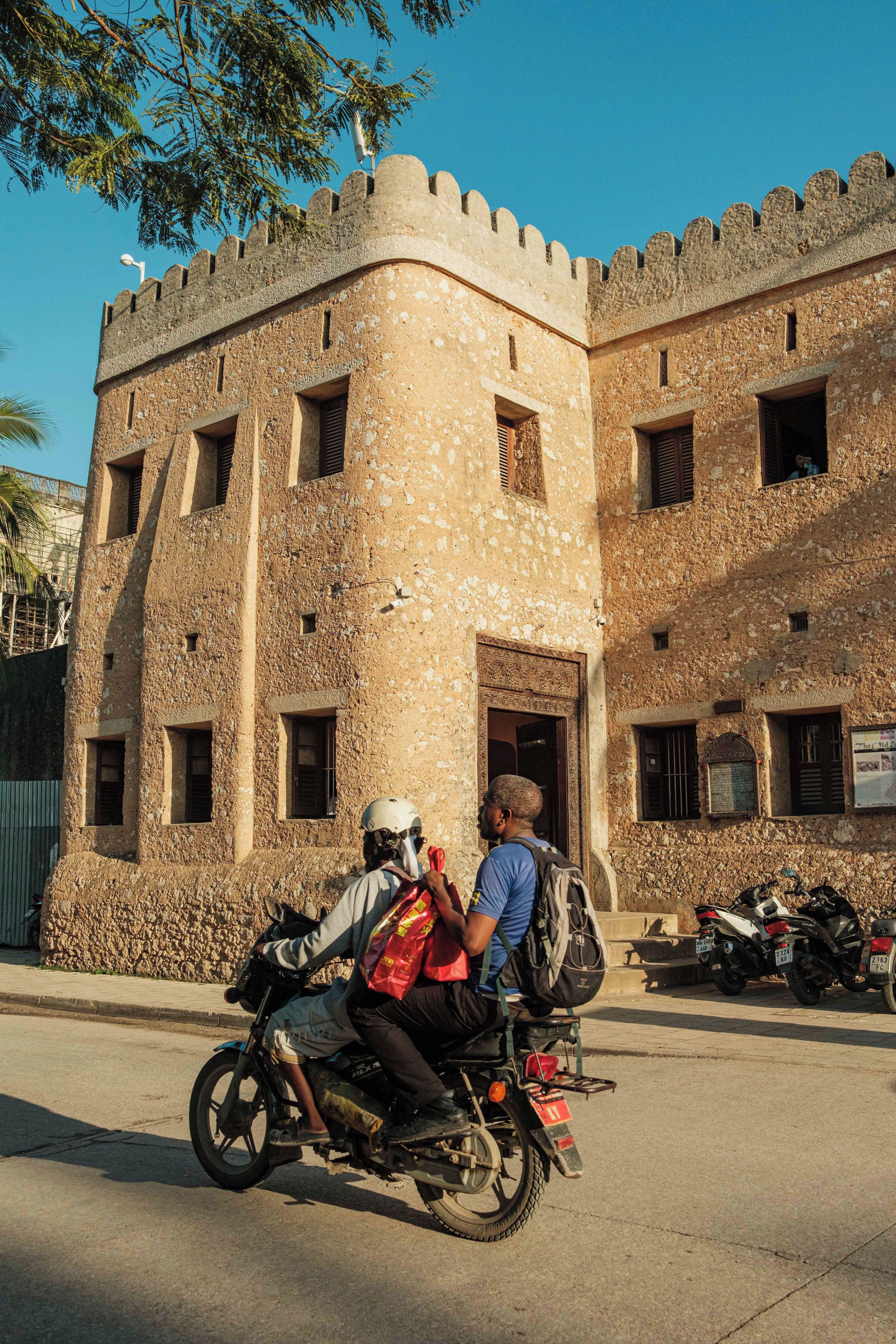Uma motocicleta com dois passageiros, um deles carregando uma bolsa vermelha, estacionada em frente a um edifício semelhante a um castelo, com várias janelas e fachada de pedra.