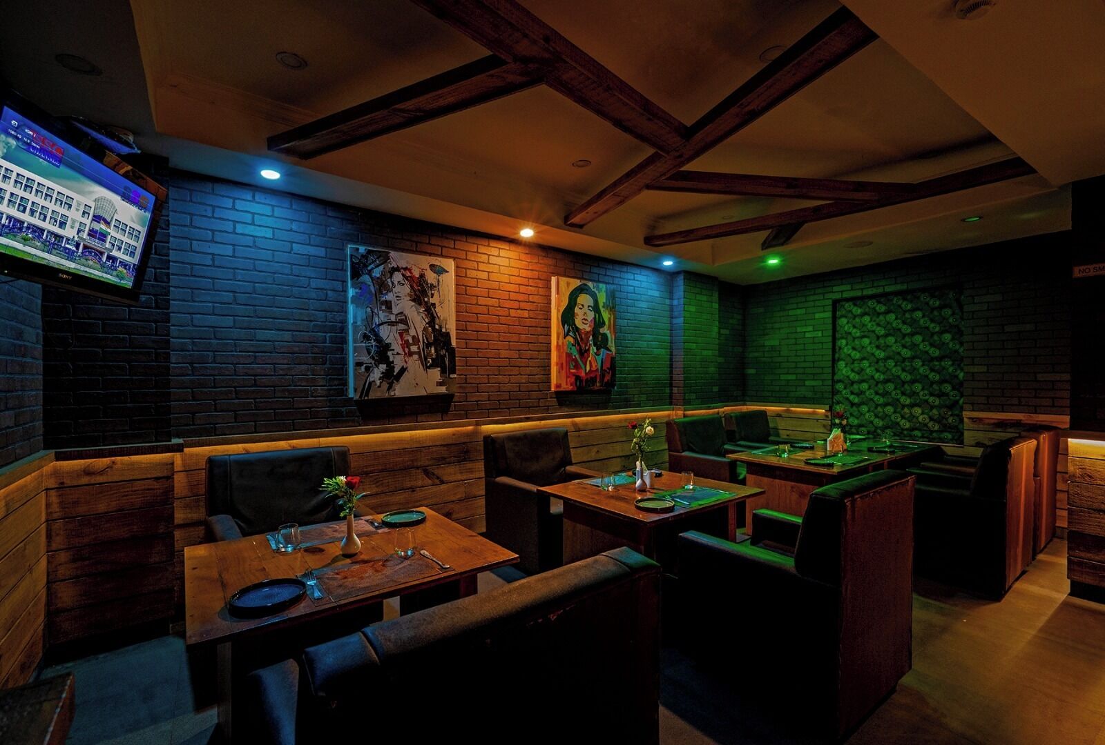 Restaurante com paredes de tijolo, vigas de madeira e iluminação ambiente verde. Mesas montadas com pratos e talheres.