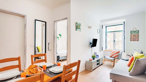 Uma sala de estar luminosa com uma mesa de jantar posta para dois, um sofá e uma televisão.